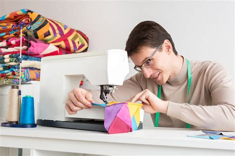 그는 조각 바느질 퀼트 및 패치워크 재단사 남성 워크샵재봉틀 웃는 재단사 남성 안경와 옷감의 밝은 패치워크의 목 스티치에 측정 테이프에서 공예 제품에 대한 스톡 사진 및