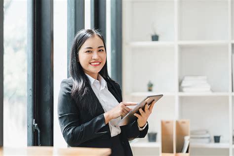 비즈니스 정장을 입은 자신감 넘치는 여성이 현대적인 사무실 환경에서 태블릿을 사용하면서 미소를 지으며 전문성과 참여를 보여줍니다 25 29세에 대한 스톡 사진 및 기타