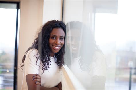 어두운 피부 긴 곱슬 머리 약 29 세의 라틴여성은 새 집의 창문너머로 반쪽 얼굴을 지지하고 사진을 위해 카메라를 바라보며 아름다운 미소를 보여줍니다 가정 생활에 대한 스톡