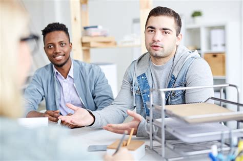 토론에 참여 하 고 사무실에서 동료에 게 그의 아이디어를 설명 하면서 몸짓 캐주얼 스웨터에 수염과 자신감 잘생긴 젊은 남자 Staff Meeting에 대한 스톡 사진 및