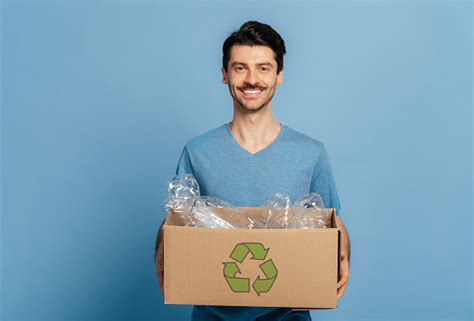 파란 티셔츠를 입은 미소짓는 양심적인 청년은 재활용을 위해 플라스틱 병으로 가득 찬 골판지 상자를 들고 카메라에 미소 짓고 고립된 파란색 배경 친환경 컨셉에 서 있습니다