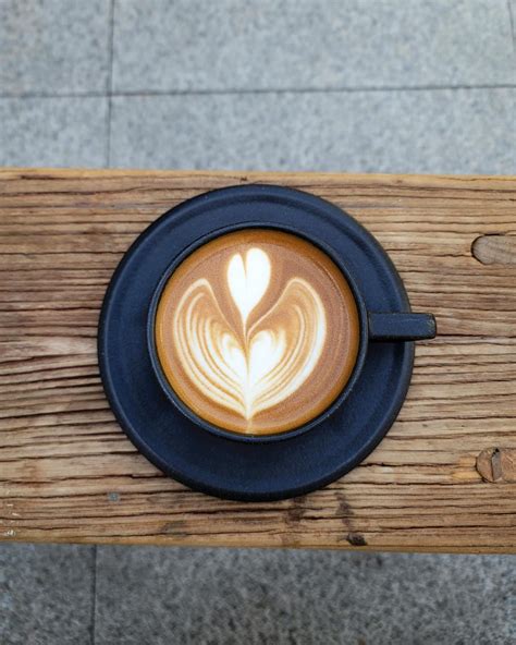 나무 테이블 위의 검은색 컵에 담긴 예술적인 라떼 · 무료 스톡 사진