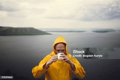 금속 머그잔을 들고 호수를 볼 수있는 비오는 날 절벽에 서있는 노란색 비옷을 입은 힙스터 여행자 방랑자와 여행 잔인한 수염 남자