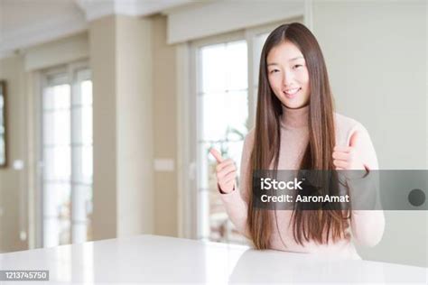 흰색 테이블에 캐주얼 스웨터를 입고 아름다운 아시아 여성은 손 엄지 손가락으로 긍정적 인 제스처를하고 성공 표지판미소와 행복