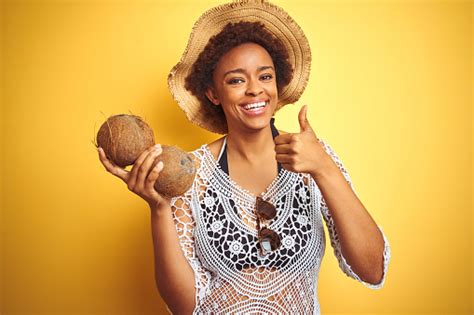 노란 고립 된 배경 위에 코코넛을 들고 있는 젊은 아프리카 계 미국인 여성은 큰 미소로 행복한 큰 미소로 확인 사인을하고 손가락으로 엄지 손가락을 위로 우수한 기호를 개념에