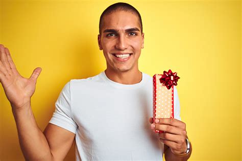 노란 고립 된 배경을 통해 생일 선물을 들고 젊은 백인 남자 매우 행복하고 흥분 큰 미소와 함께 비명을 지르는 승리를 축하하는 우승자 표정 공휴일에 대한 스톡 사진 및 기타