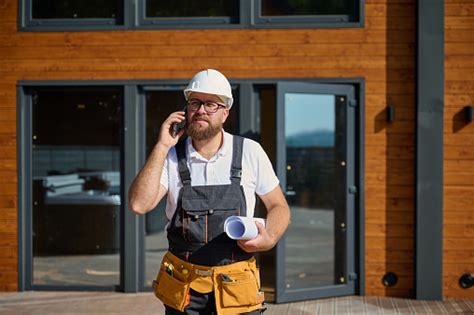 건축가는 스마트폰으로 계획을 잡고 말합니다 턱수염을 쓴 건축가 프로젝트 청사진 종이 계획 프로젝트 청사진을 들고 건설 현장을 바라 보는 새로운 현대 주택의 Comlex
