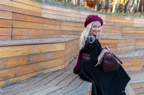 쾌활한 한 여성이 가을 푸른 하늘을 배경으로 카메라를 바라보며 부르고뉴 코트와 버렛을 입고 나무 여름 극장 벤치에 앉아 있다 공원에 대한 스톡 사진 및 기타 이미지 Istock