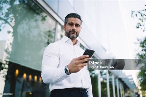 공식적으로 옷을 입은 사업가 휴대 전화와 금융 지구의 도시 환경에 서서 전화 기술을 사용하는 동안 카메라를보고 스마트 폰으로 성숙한 백인 고용주의 절반 길이 35 39세에