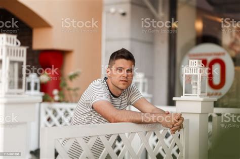 세련된 헤어 스타일과 세련된 줄무늬 티셔츠에 좋은 매력적인 젊은 남자는 여름 카페의 테라스에 나무 흰색 울타리 근처에 휴식된다