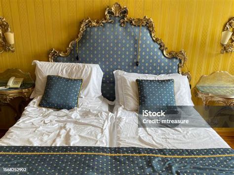 화려한 프레임 무늬가 있는 패딩 처리된 헤드보드 흰색 이불 커버 파란색과 금색 줄무늬 쿠션 벽걸이형 전기 램프 전화기 야간 스탠드 전경에 초점을 맞춘 흰색 호텔 베개 더미가