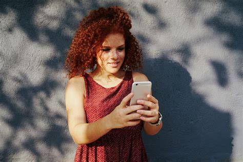 빨간 드레스를 입고 얼굴에 주 근 깨와 귀여운 빨간 머리 여자 학생이 하고있다 Selfie 스마트폰 카메라 화창한 날 야외에서 회색 배경에 서 있는 동안 사진의 허리 5g에