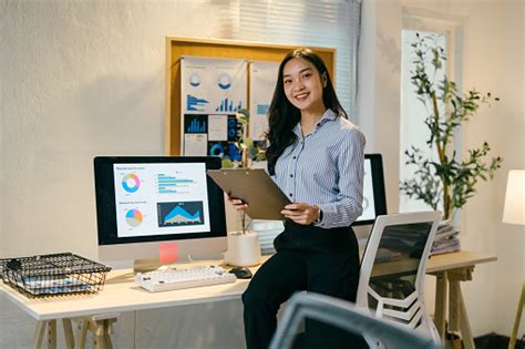 젊은 아시아 사업가가 현대 사무실에 서서 클립 보드를 들고 카메라 앞에서 미소 짓고 차트와 그래프를 컴퓨터 화면과 코르크 보드에 표시하고 있습니다 20 29세에 대한 스톡