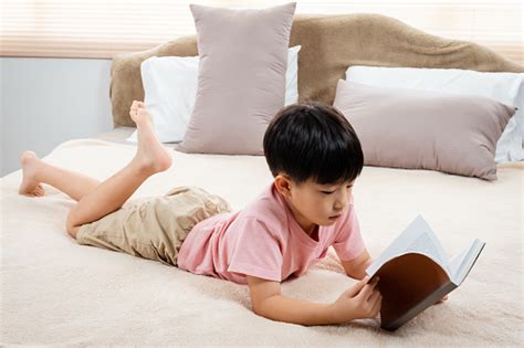 분홍색 셔츠를 입은 아시아 소년은 편안한 자세로 방의 침대에 평평하게 누워 있으며 좋아하는 책을 들고 손에 책의 그림을 기쁜 마음으로 열었습니다 8 9 살에 대한 스톡 사진