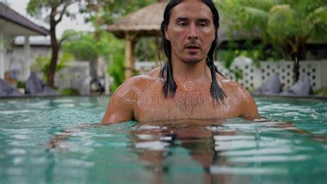 A Handsome Man With A Naked Torso And Long Hair Stands In A Waterfall Into The Water Slow