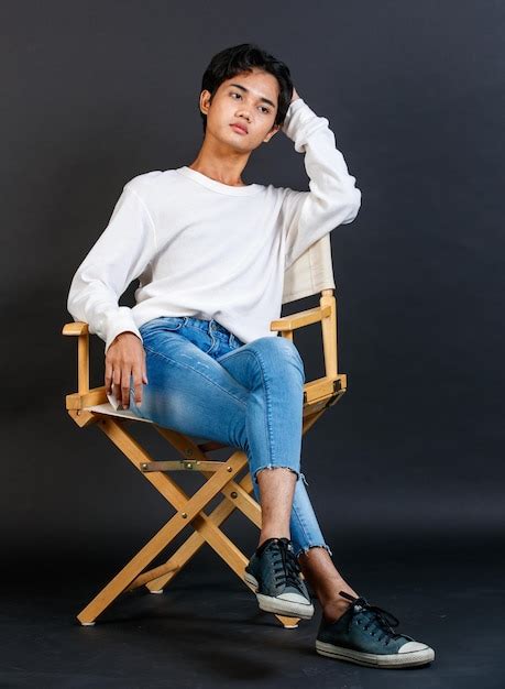 검은 배경에 몸짓으로 포즈를 취하는 나무 의자에 다리를 꼬고 앉아 캐주얼 복장을 한 아시아 젊은 Lgbtq 게이 글래머 잘생긴