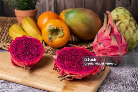 피타 하야 또는 용 과일은 나무 판에 두 개로 나뉘어 분홍색 내부와 검은 씨앗을 드러내고 배경에는 체리 모야 망고 감 바나나와 같은 열대 과일이 전경에 있습니다 0명에 대한