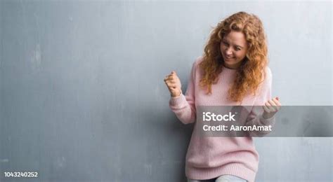 젊은 빨강 머리 여자 회색 그런 지 벽 입고 위에 분홍색 스웨터 매우 행복 하 고 팔 웃 고 성공을 위한 비명 우승자 제스처를 하 고 흥분 축 하 개념입니다 곱슬 머리에