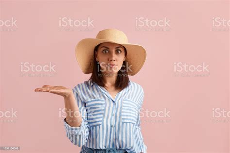 어두운 머리를 가진 예쁜 여자는 손바닥을 보여줍니다 할인 또는 좋은 거래를 보여줍니다 카메라를 똑바로 보인다 밀짚 모자를 입고 줄무늬 셔츠 벨트 청바지 핑크 배경에 여름