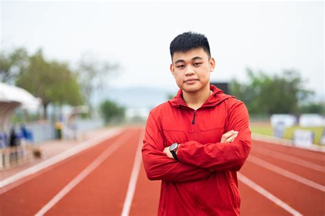 스포츠웨어 빨간 재킷을 입은 젊은 잘 생긴 아시아 남자는 트랙 스타디움에서 야외로 달리는 포스트를 서있다 길에서 조깅하는 중국 남자의 초상화 운동 선수 야외 개념 훈련 30