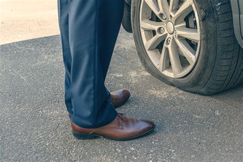 파란색 양복을 입은 사업가가 차 근처에 서서 바퀴에 구멍이 뚫린 정도정도를 확인한다 타이어에 구멍 개념 견인차에 대한 스톡 사진