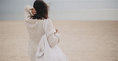 바람이 부는 머리와 토트백을 들고 모래 해변에서 바다로 걸어가는 아름다운 세련된 여성 평온한 순간 니트 스웨터를 입은 세련된