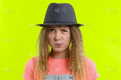 一个漂亮有趣的十几岁女孩的肖像与沉思的脸，金发女孩卷头发在帽子与深思的表情，压在一起的嘴唇，黄色的工