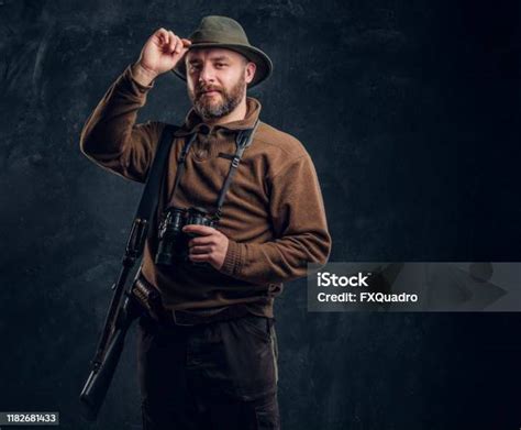 소총과 쌍안경을 쓴 수염난 사냥꾼이 모자를 수정하고 카메라를 바라보고 있습니다 어두운 벽 배경에 대한 스튜디오 사진 Championship에 대한 스톡 사진 및 기타 이미지