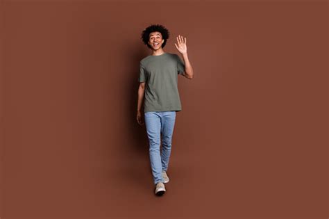 회색 티셔츠와 청바지에 물결 모양의 머리를 가진 젊은이 남자의 전신 길이 사진 산책 웨이브 손 환영 갈색 배경에 고립 된 20 24세에 대한 스톡 사진 및 기타 이미지