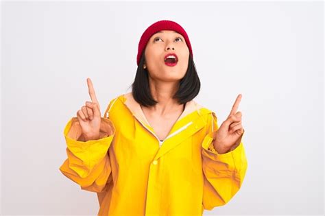 고립 된 흰색 배경 위에 비옷과 양모 모자를 쓰고 있는 젊은 아름다운 중국 여성은 손가락과 팔을 들고 위를 올려다 보고 놀랐습니다 가리키기에 대한 스톡 사진 및 기타 이미지