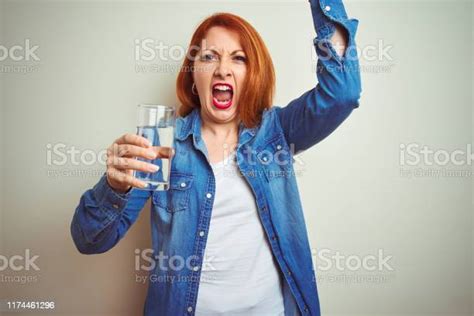 흰색 고립 된 배경을 통해 물 잔을 마시는 젊은 아름다운 빨간 머리 여자는 짜증과 분노와 함께 고함 미친 고함 제기 손으로 소리 분노 개념 기이함에 대한 스톡 사진 및 기타