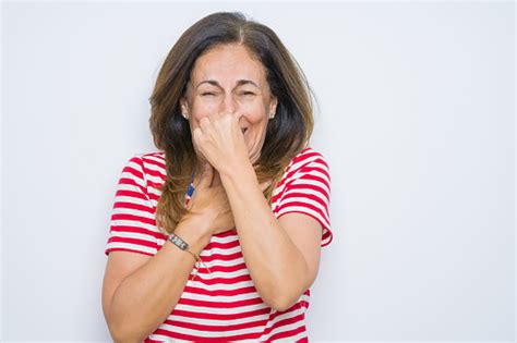 흰색 고립 된 배경 위에 서있는 중년 여성은 냄새가 없고 역겨운 냄새 참을 수없는 냄새를 맡고 코에 손가락으로 숨을 들고 있습니다 나쁜 냄새 개념 T 셔츠에 대한 스톡 사진