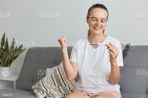 임신 검사를 손에 들고 흰색 티셔츠를 입고 긍정적 이거나 부정적인 결과를 기뻐하며 집에서 소파에 앉아 주먹을 움켜 쥐는 매우