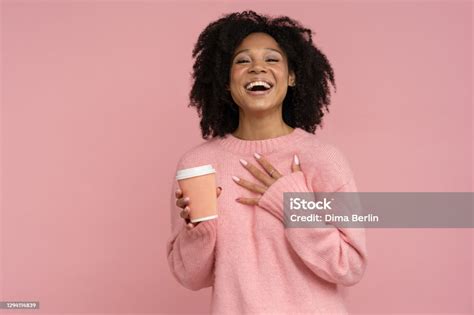 곱슬 머리를 가진 쾌활한 어두운 피부 밀레니엄 여성은 뜨거운 커피 또는 차 테이크 아웃의 종이 컵을 들고 카메라를보고 핑크 스웨터를 착용합니다 스튜디오 파스텔 핑크 배경