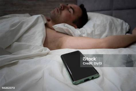 그가 자는 동안 핸드폰을 들고 있는 스마트폰 사용자 침대에서 졸고 있는 청년 모바일 중독 개념 텍스트와 디자인을 위한 빈 스마트폰 화면 공간을 복사합니다 알람시계에 대한