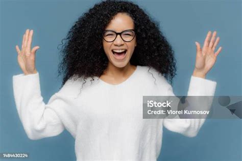 곱슬 머리와 혼합 레이스 여자를 기 뻐 손을 제기 긍정적 인 감정에서는 흰색 스웨터를 입고 입을 열어 유지 파란색 배경에 대 한 의미 얼굴 표정 개념 20 24세에 대한