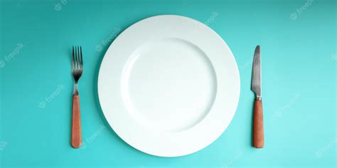 Белая пустая тарелка с вилкой и ножом лежала на фоне синего стола ...
