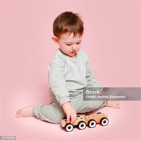 행복한 유아 아기는 분홍색 배경에 장난감 자동차를 가지고 놀고 있다 어린 소년은 나무 장난감 자동차를 굴립니다 아이 나이 1 년 8 개월 전체 신장 건강한 생활방식에 대한