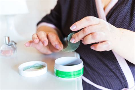 목욕 가운을 입은 노인 여성은 녹색 알로에 눈 패치를 사용하여 얼굴 화장품을 적용합니다 자기 관리 노화 피부를 위한 트리트먼트 여성의 아늑한 나무 침실 서랍의 가슴 거울