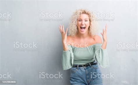 그런 지에 곱슬 머리 젊은 금발의 여 자가 회색 배경 축 하 미친 고 제기 및 오픈 눈 비명 흥분 하는 무기와 성공을 위한 깜짝 놀라게 우승자 개념 곱슬 머리에 대한 스톡