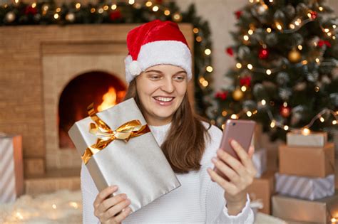그녀의 휴대 전화와 선물 상자를 손에 들고 매력적인 귀여운 아름다운 매력적인 소녀의 사진 미소 여성은 장식 된 거실에서 포즈 그녀의 크리스마스 선물과 화상 통화 또는 셀카를