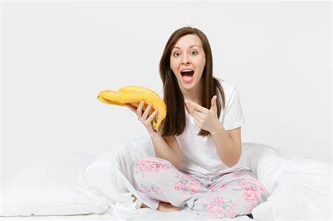 행복 한 젊은 여 자가 하얀 시트 베개 담요 흰색 바탕에 배치와 함께 침대에 앉아 재미 뷰티 여자 방에서 시간을 보내는 바나나를 잔뜩 들고 휴식 휴식 좋은 분위기 개념