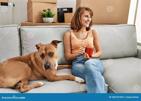 молодая кавказская женщина пьёт кофе сидя на диване с собакой дома Стоковое Изображение
