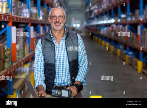 Senior Male Warehouse Worker Leaning On Rolling Cart Holding Laptop And