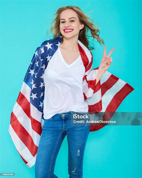 나는 미국을 사랑 해요 행복 한 젊은 웃는 여자 청바지와 흰색 티셔츠에 평화 서명 만들기 및 파스텔 파란색 배경에 서 있는 동안 카메라를 보고 미국 국기에 싸여 있다