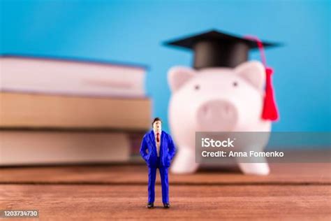 미니어디어 사람들 졸업 모자를 쓰고 연한 파란색 백라운드에 많은 책을 들고 돼지 저금통 앞에 서 있는 남자 비용 절감 금융