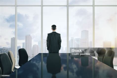 현대적인 회의실 내부에 서있는 젊은 사업가의 뒷모습은 테이블 의자 및 하늘 구름 전망과 일광이있는 탁 트인 창문에 반사되어 있습니다 Ceo 및 컨퍼런스 개념 회의실에 대한