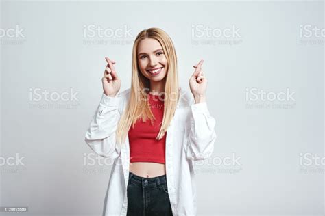 행운을 기대 합니다 회색 배경에 서 있는 동안 손가락이 교차 잡고 흰색 셔츠에 젊은 금발 여자를 웃 고 Wishness 개념입니다 가능성에 대한 스톡 사진 및 기타 이미지