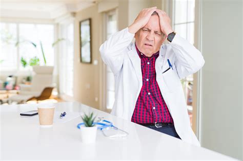 의료 코트를 쓰고 있는 잘생긴 노인 닥터는 두통으로 고생 하는 클리닉에서 고 통과 편두통 때문에 스트레스를 받고 있습니다 머리에 손을 걱정하는에 대한 스톡 사진 및 기타