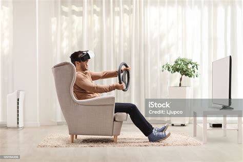 가상 현실 헤드셋을 사용하여 안락 의자에 앉아 스티어링 휠을 들고 있는 청년 운전하기에 대한 스톡 사진 및 기타 이미지